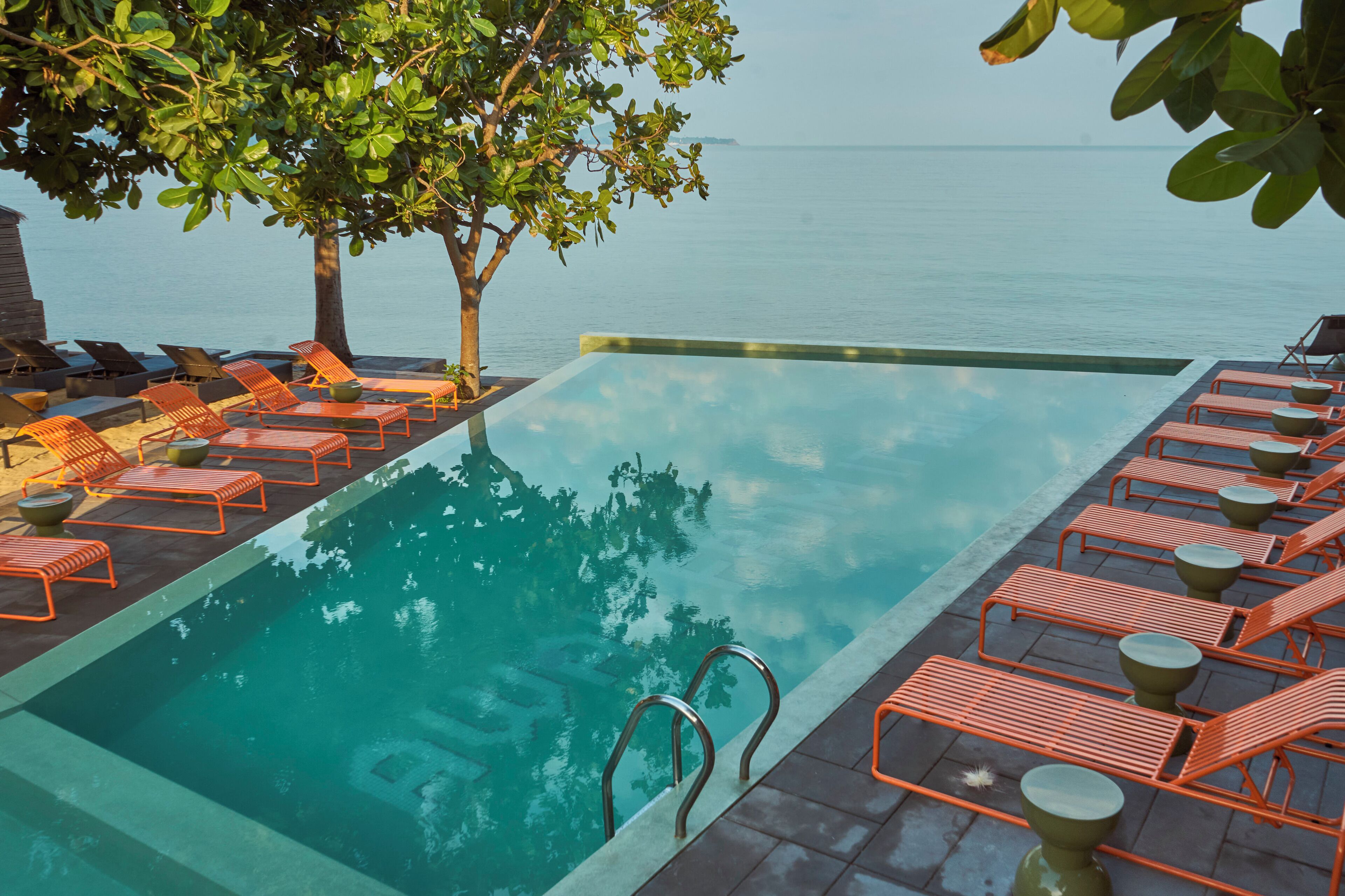 Piscina com vista clara para o mar, rodeada por espreguiçadeiras laranja e uma árvore.