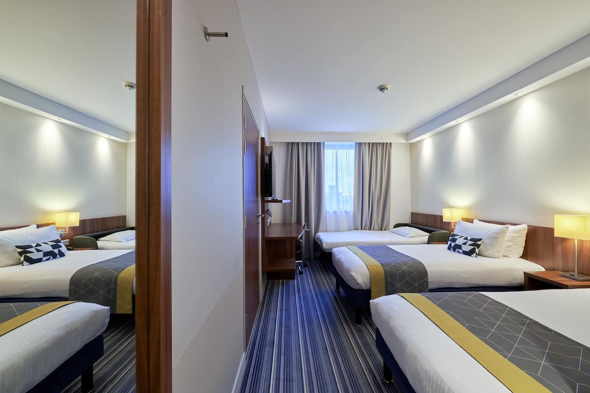 Quarto de hotel com duas camas, uma cabeceira de madeira, uma escrivaninha e uma janela com cortinas.