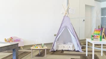 Uma sala de jogos infantil com uma pequena mesa, um carrinho de brinquedo, um urso de pelúcia e uma tenda.