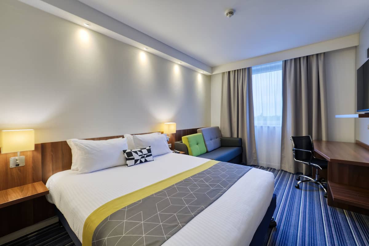 Quarto de hotel com cama grande, escrivaninha, cadeira, luminária e janela com cortinas.