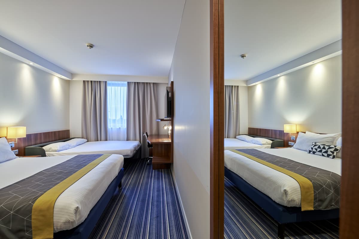 Quarto de hotel com duas camas, uma escrivaninha, uma cadeira, televisão e janela com cortinas.