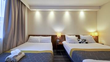 Quarto de hotel com duas camas, uma mesa de cabeceira com abajur e uma janela com cortinas.