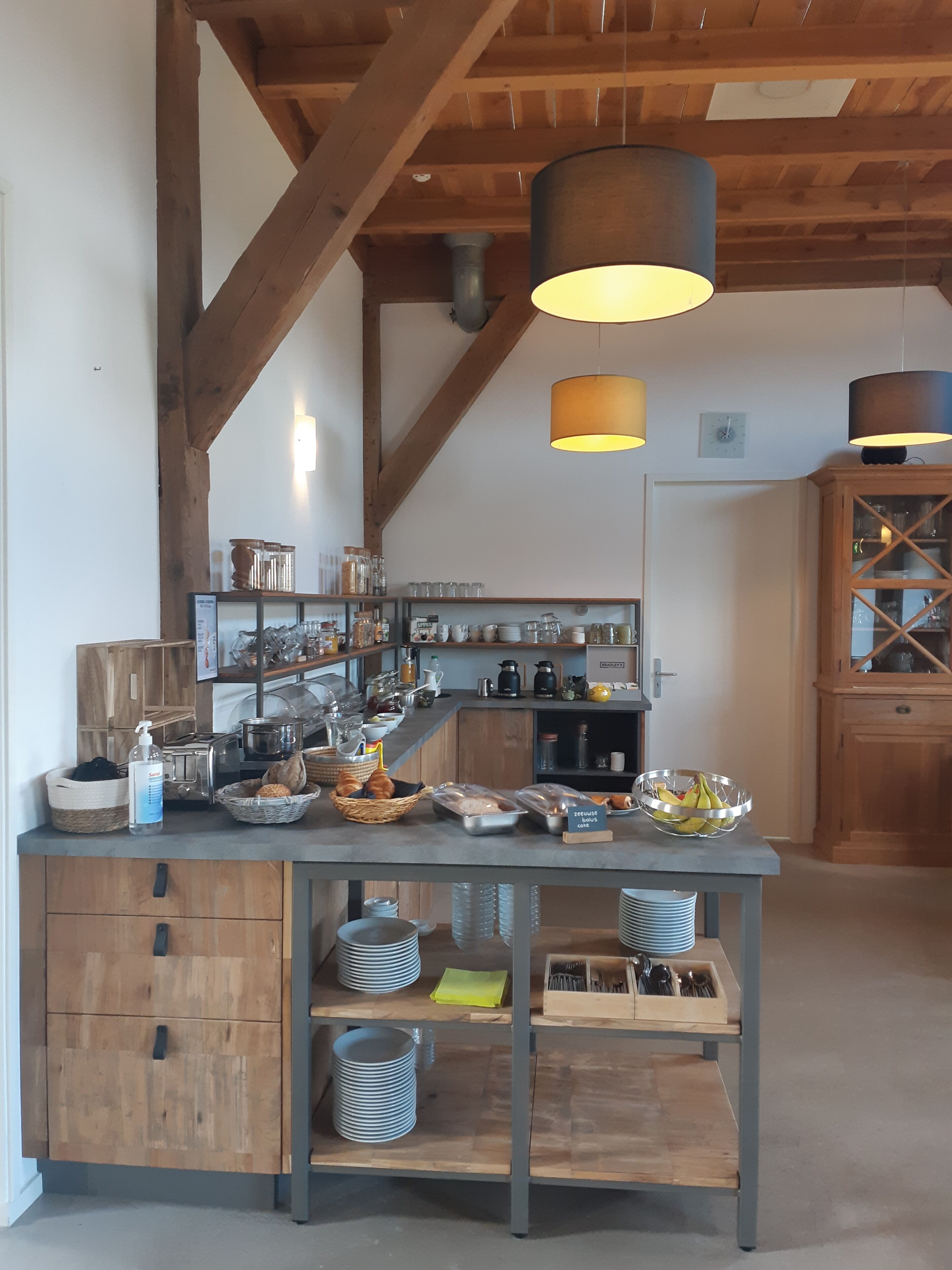 Cozinha com vigas de madeira, ilha central com diversos alimentos e luminárias pendentes.