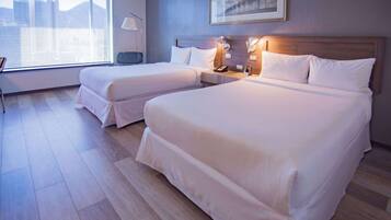 Quarto de hotel com duas camas, vista para a cidade, um quadro pendurado na parede e uma mesa de cabeceira com abajur.