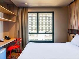 Quarto de hotel com cama, secretária com telefone, cadeira e janela com vista para edifícios.