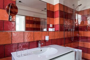 Uma casa de banho moderna com uma pia branca, paredes revestidas de azulejos vermelhos, cabine de duche em vidro e um espelho grande.