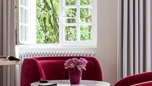 Quarto com uma janela grande, sofá vermelho, mesa redonda pequena com um vaso de flores e piso xadrez.