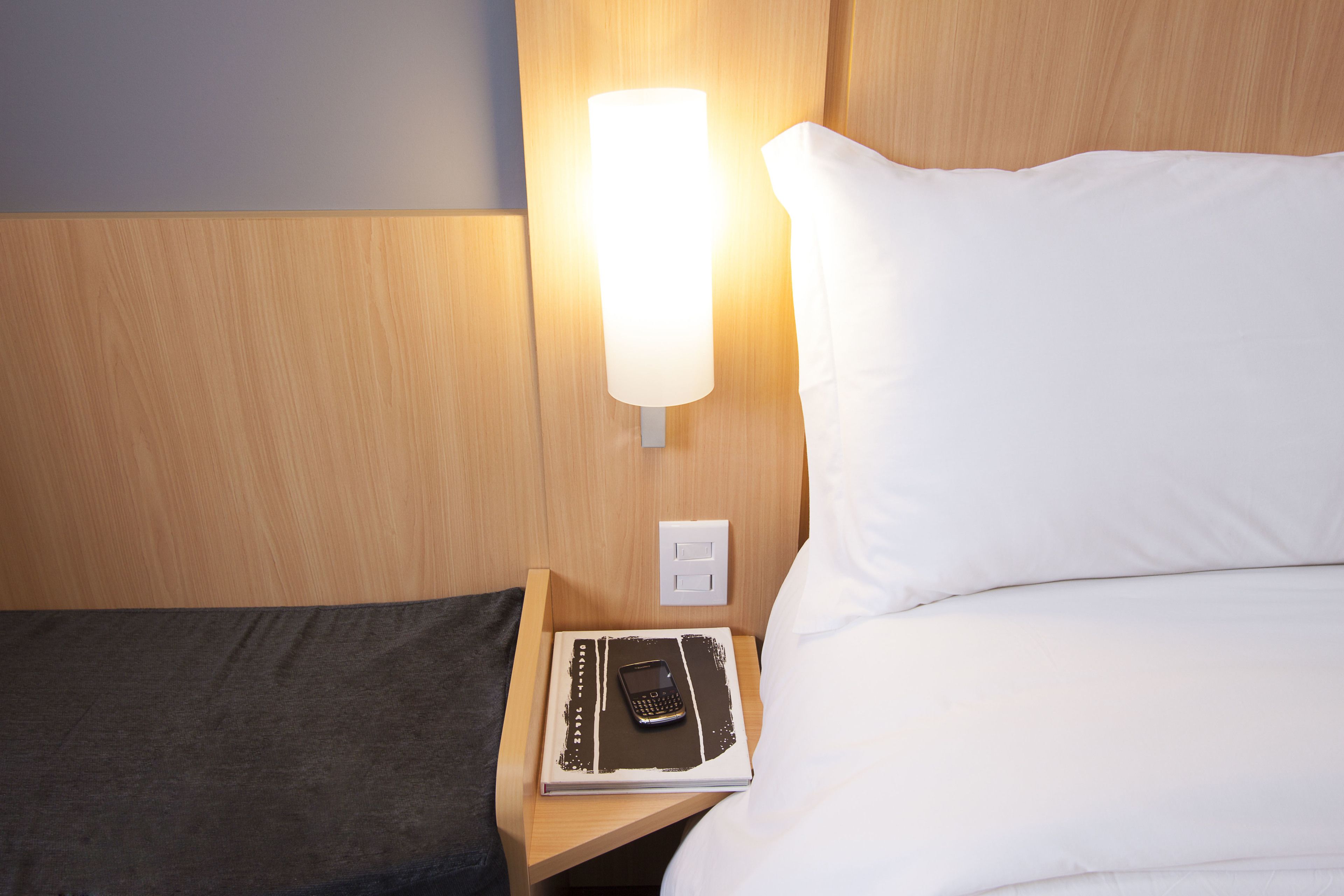 Cama bem arrumada com lençóis e almofadas brancos, mesa de cabeceira com abajur e um livro com um smartphone em cima.