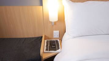 Cama bem arrumada com lençóis e almofadas brancos, mesa de cabeceira com abajur e um livro com um smartphone em cima.