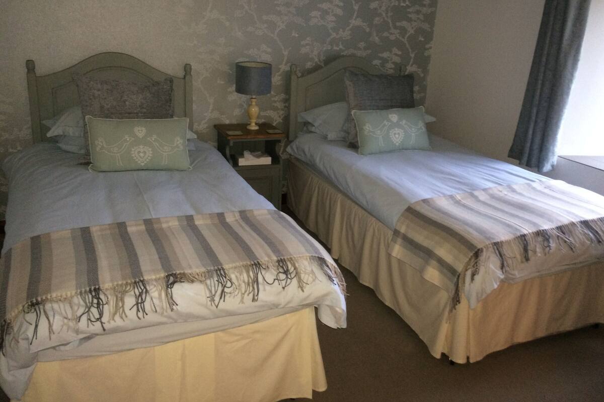 Duas camas de solteiro com roupa de cama listrada, uma mesa de cabeceira com abajur e um papel de parede estampado com flores.