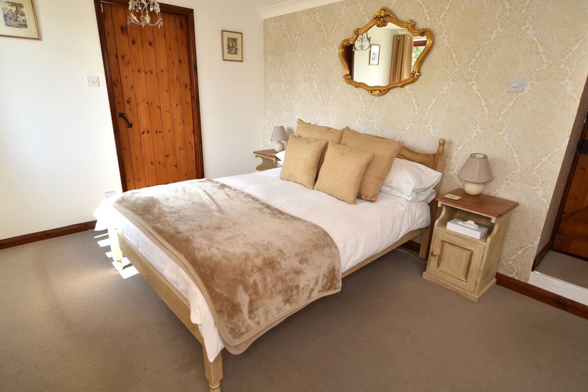 Quarto com cama de madeira, roupa de cama bege, mesa de cabeceira com abajur, espelho e porta.