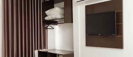 Quarto de hotel com televisão de tela plana fixada na parede, ar-condicionado embutido, guarda-roupa com prateleira suspensa e cortina.