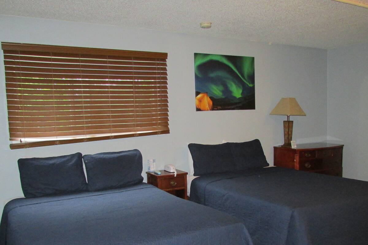 Quarto com duas camas, janela com persianas, pintura da aurora boreal, luminária e mesa de cabeceira.