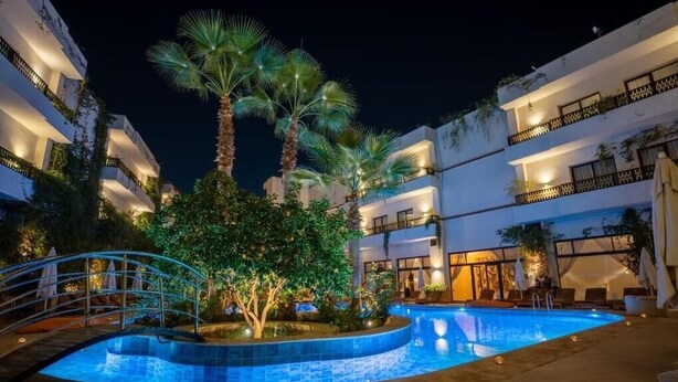 Vista noturna de um pátio de hotel com piscina, palmeiras e várias varandas.