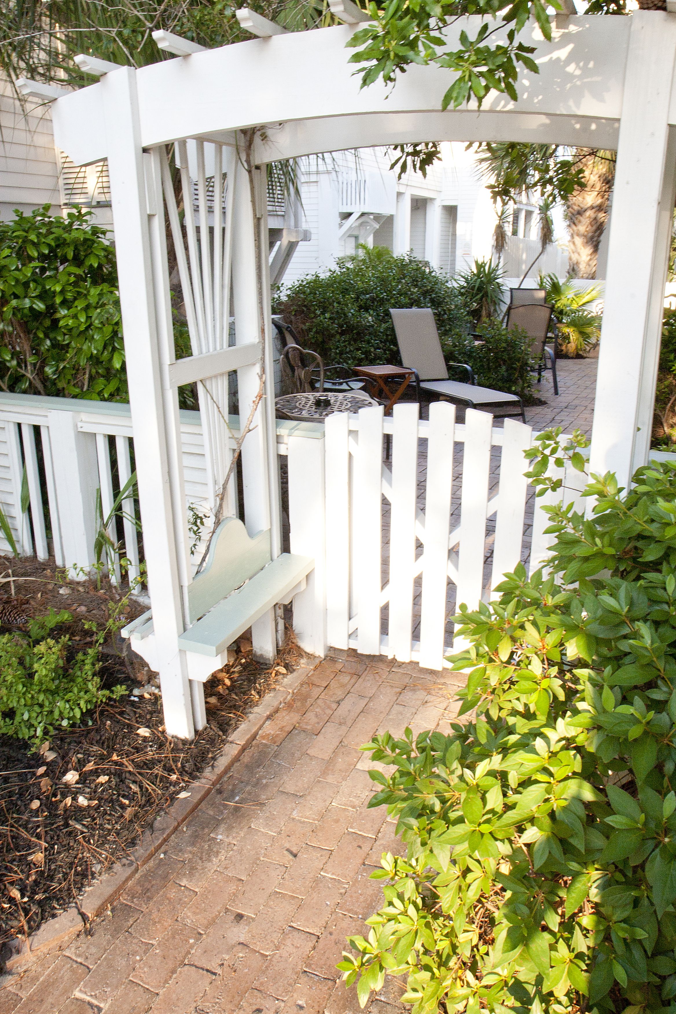 Uma pérgola de madeira branca com um banco, cercada por vegetação e um cerca de piquetes brancos.