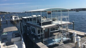 Um barco atracado num cais, com área de estar coberta e vista para a água e edifícios ao longe.