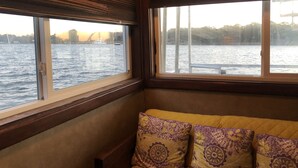 Uma cabana com vista para um rio e uma ponte. Há uma janela com persianas, um banco acolchoado e almofadas estampadas.