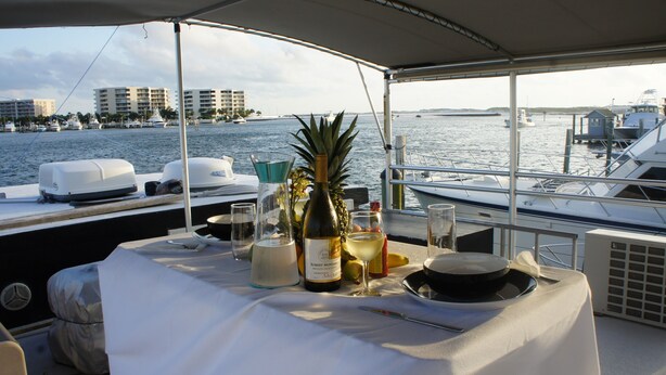 Mesa posta para refeição com uma garrafa de vinho, uma abacaxi e copos em um barco, com vista para a marina e edifícios ao fundo.