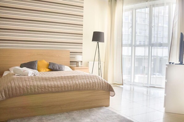 Quarto moderno com uma cama grande, uma mesa de cabeceira, uma luminária e vista da cidade através de grandes janelas.
