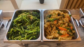 Uma mesa de buffet com duas bandejas de metal, uma com vegetais verdes e outra com macarrão e vegetais.