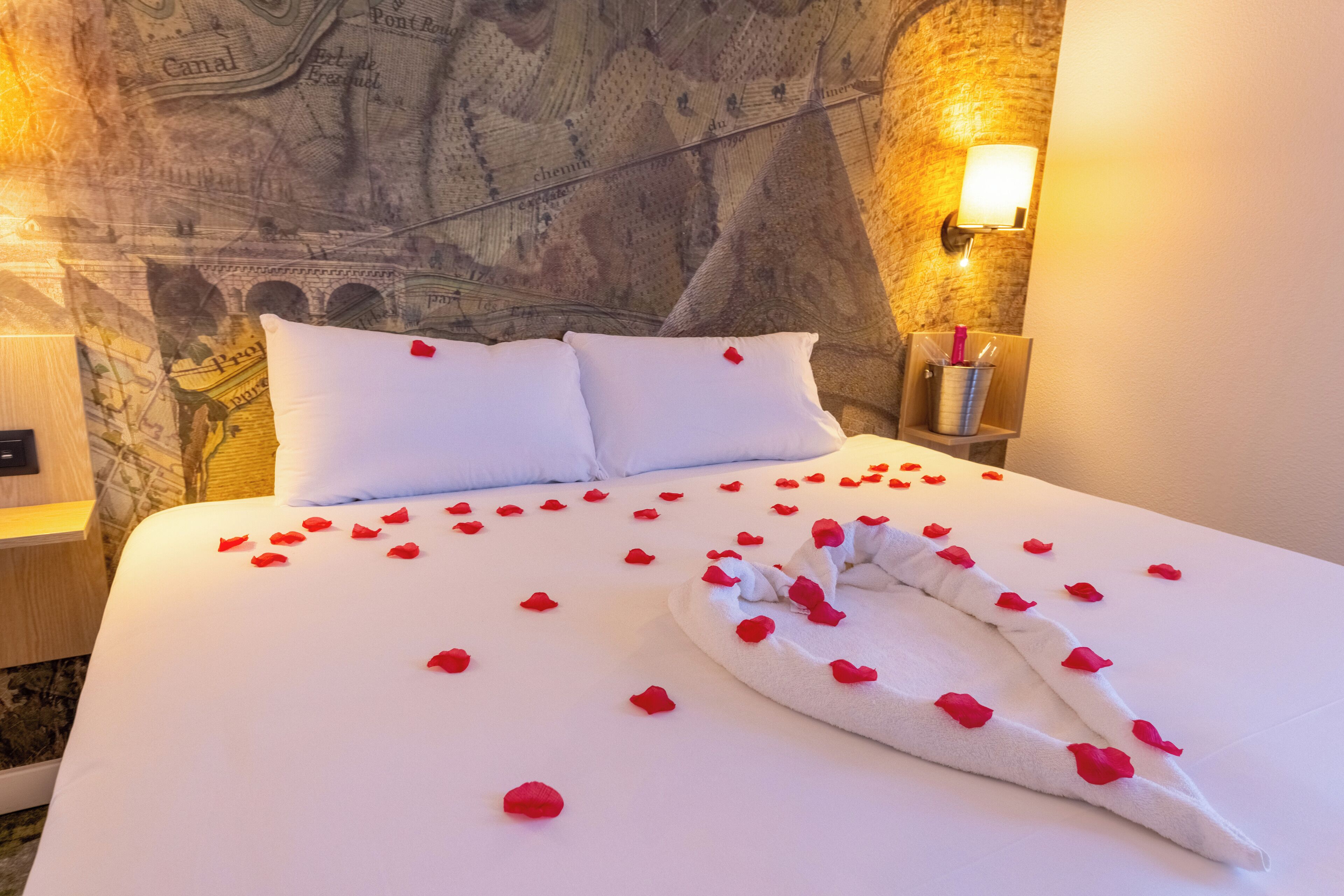 Cama bem arrumada com roupa de cama branca e pétalas de rosa dispostas em formato de coração.
