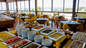 Disposição de buffet com diversos alimentos e vista para o oceano através de grandes janelas.
