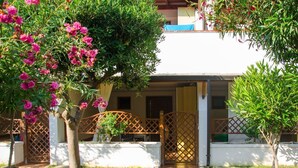 Um edifício com fachada branca, um portão de madeira e um pequeno jardim com flores cor-de-rosa e arbustos verdes.