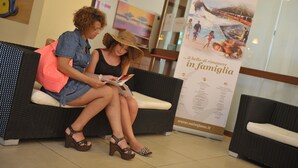Duas pessoas sentadas num sofá de vime em um hall de entrada, com um cartaz a anunciar Saint Jane Hotels & Suites.