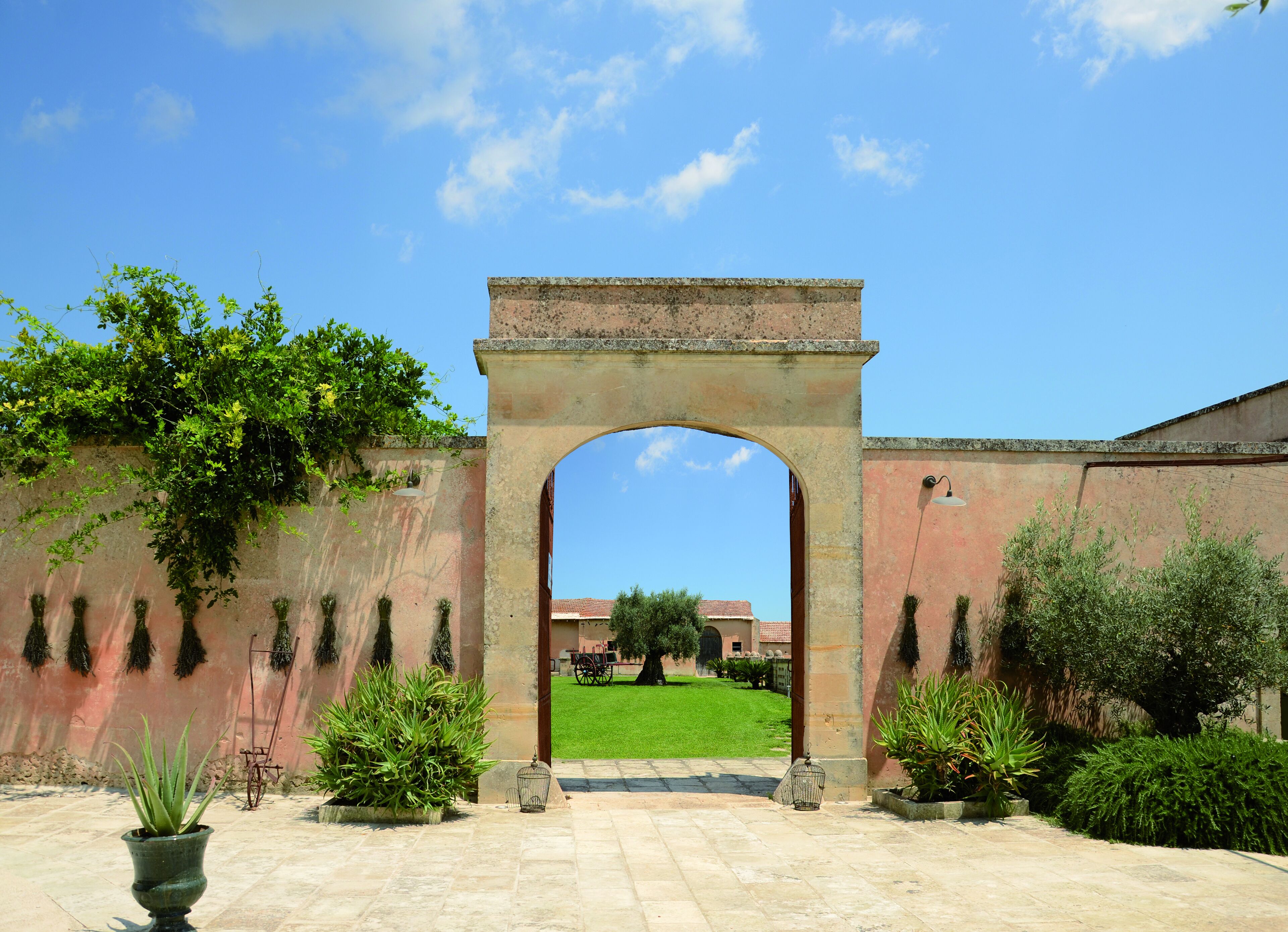 Uma entrada em arco para um edifício, com um pátio pavimentado, rodeado por vegetação e um céu azul límpido.