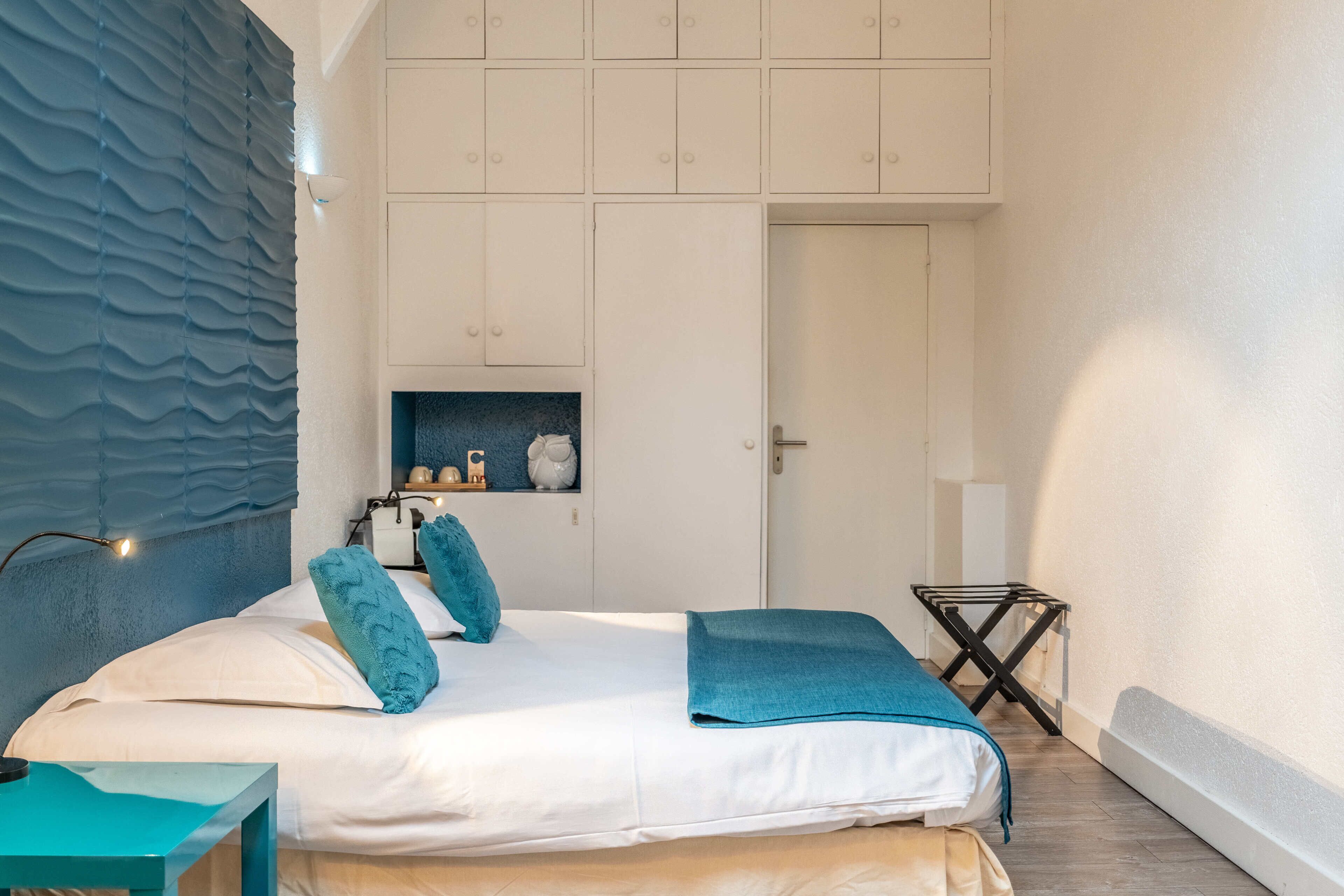Quarto com cama, mesa de cabeceira, prateleira na parede com objetos decorativos e porta que dá para outro cômodo.