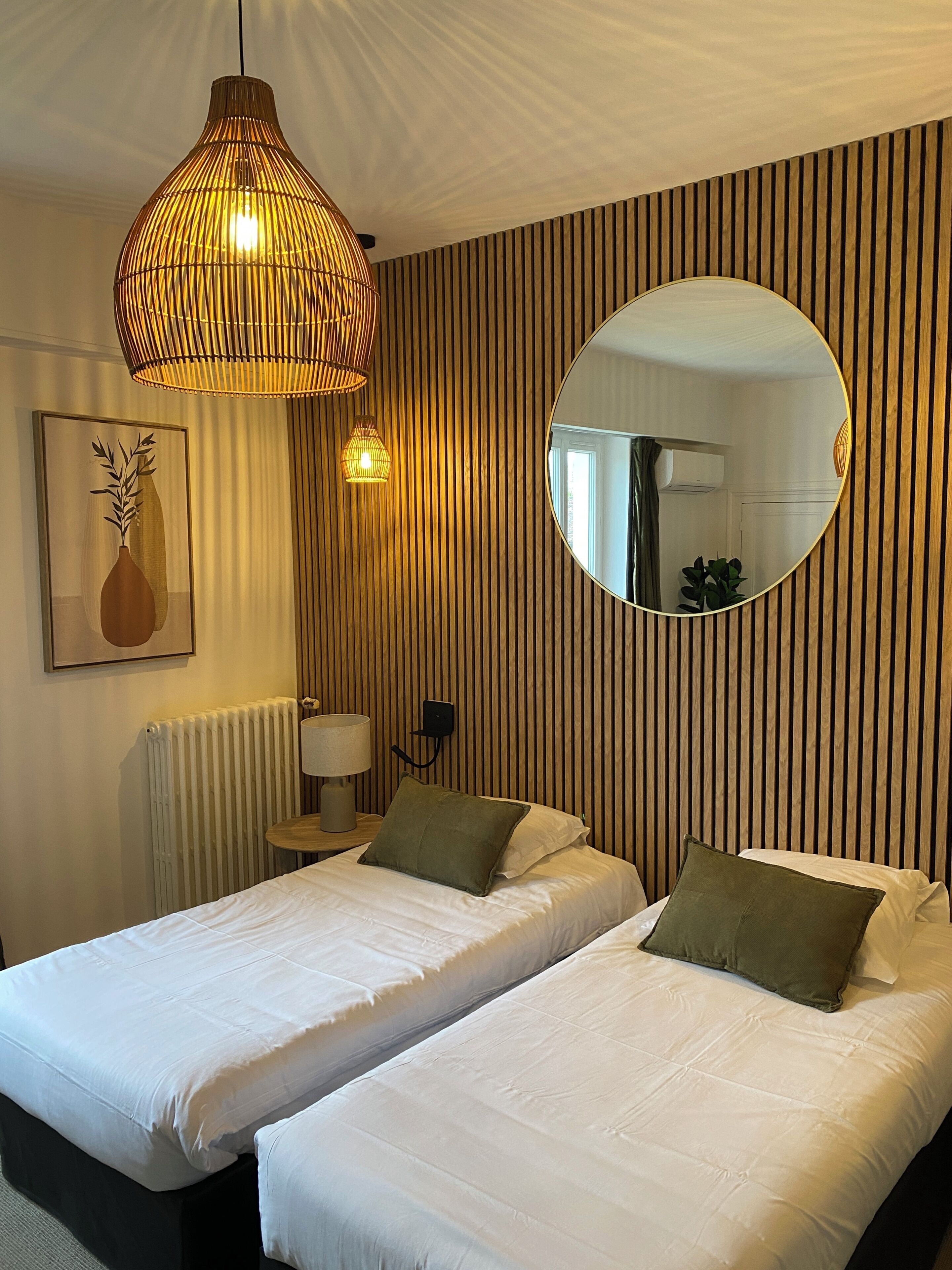 Quarto de hotel com duas camas, um espelho redondo, uma luminária pendente e um radiador.