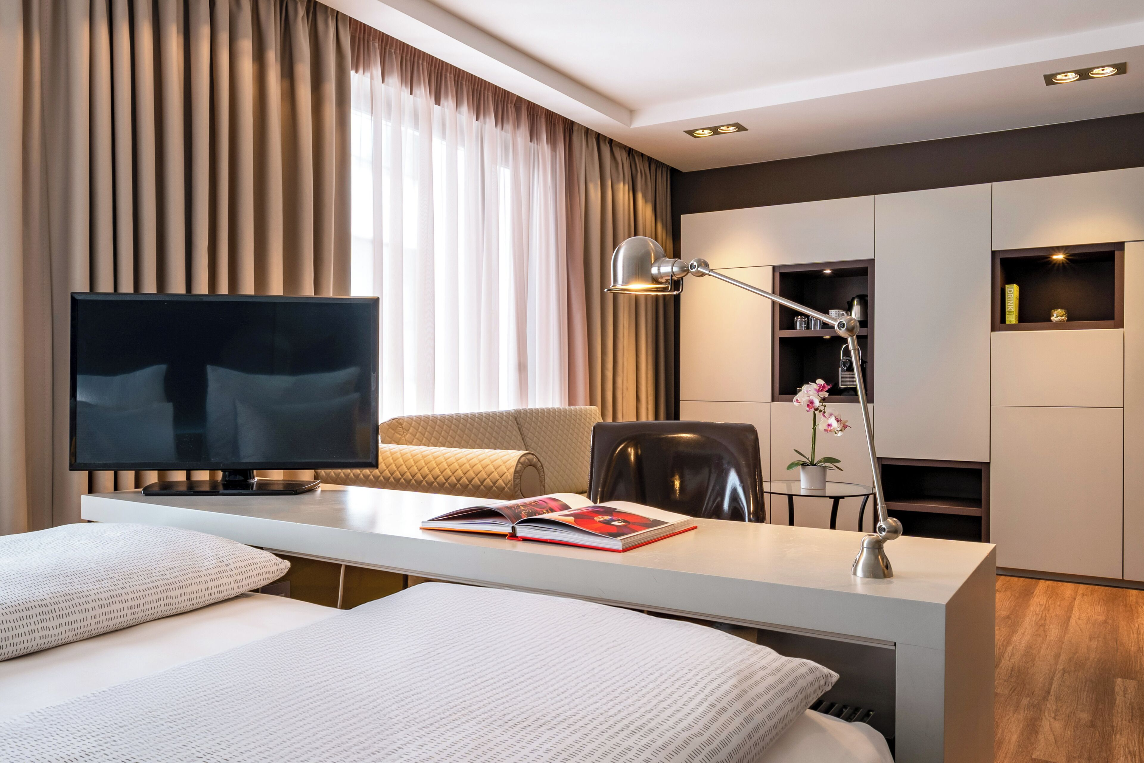 Quarto de hotel moderno com televisão de tela plana, escrivaninha com abajur, cadeira e cama com almofadas.