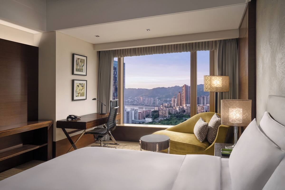 Quarto de hotel com cama, escrivaninha, cadeira, sofá e uma grande janela com vista para a cidade.