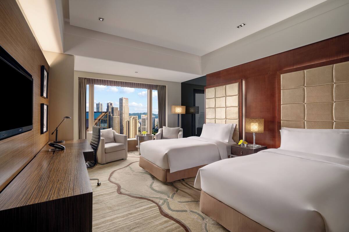 Quarto de hotel com duas camas, uma televisão, uma escrivaninha e vista para a cidade.