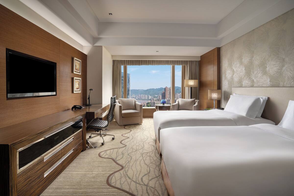 Quarto de hotel com uma televisão grande, duas camas, uma escrivaninha e vista para a cidade.