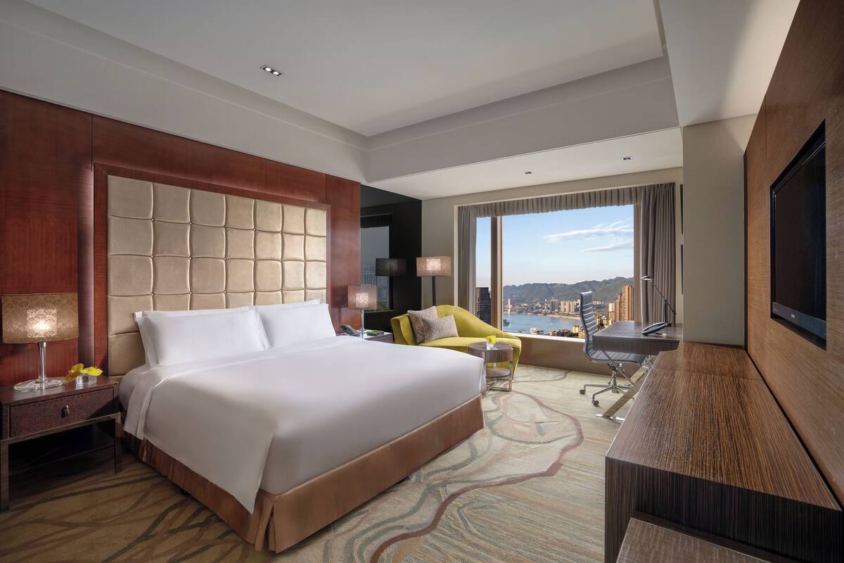 Quarto de hotel com uma cama grande, uma escrivaninha, uma cadeira, uma televisão e vista para o horizonte da cidade.