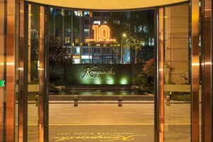Entrada de hotel com um grande letreiro iluminado com o número '10', um elemento decorativo de água e uma placa indicando 'Kempinski Hotels Desde 1897'.