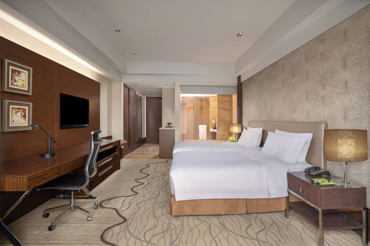 Quarto de hotel com cama, escrivaninha, cadeira, televisão e mesa de cabeceira com abajur.