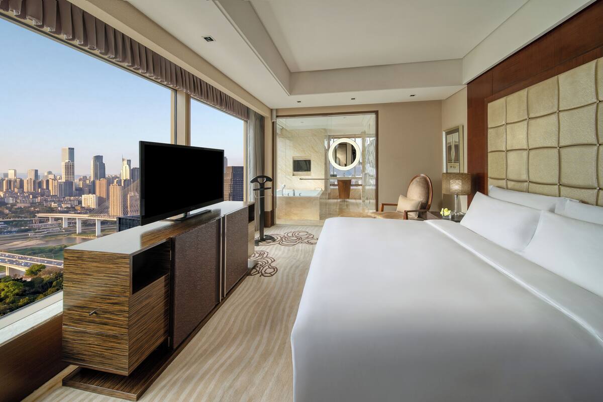 Quarto de hotel com uma cama grande, televisão em um console de madeira, vista para a cidade, banheiro com espelho e mesa de cabeceira com abajur.