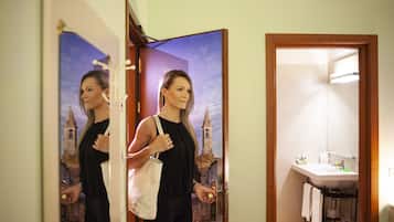 Quarto de hotel com um espelho grande refletindo uma mulher segurando um telefone e uma bolsa branca. O quarto possui um banheiro com pia e porta-toalhas.