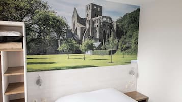 Quarto de hotel com uma cama, mesa de cabeceira de madeira, parede branca e um pôster grande de um castelo.