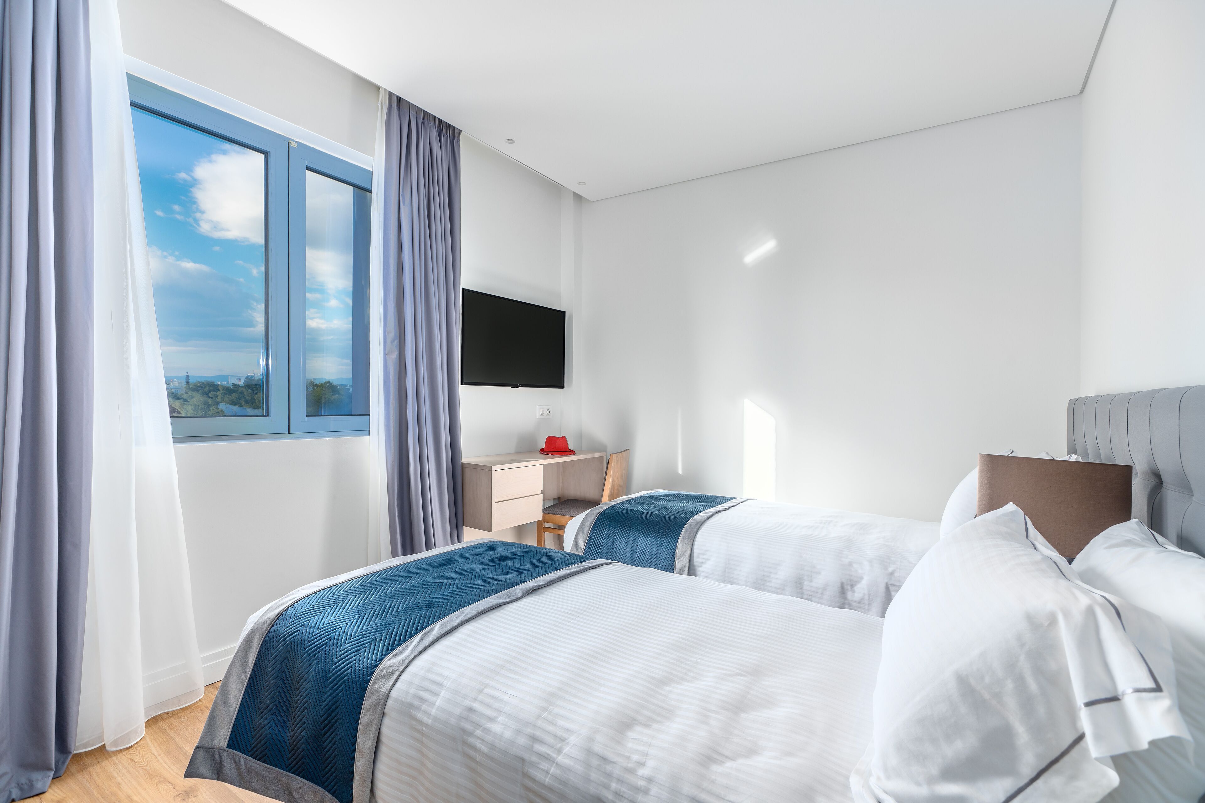 Quarto de hotel moderno com duas camas, uma escrivaninha, televisão e janela com cortinas.