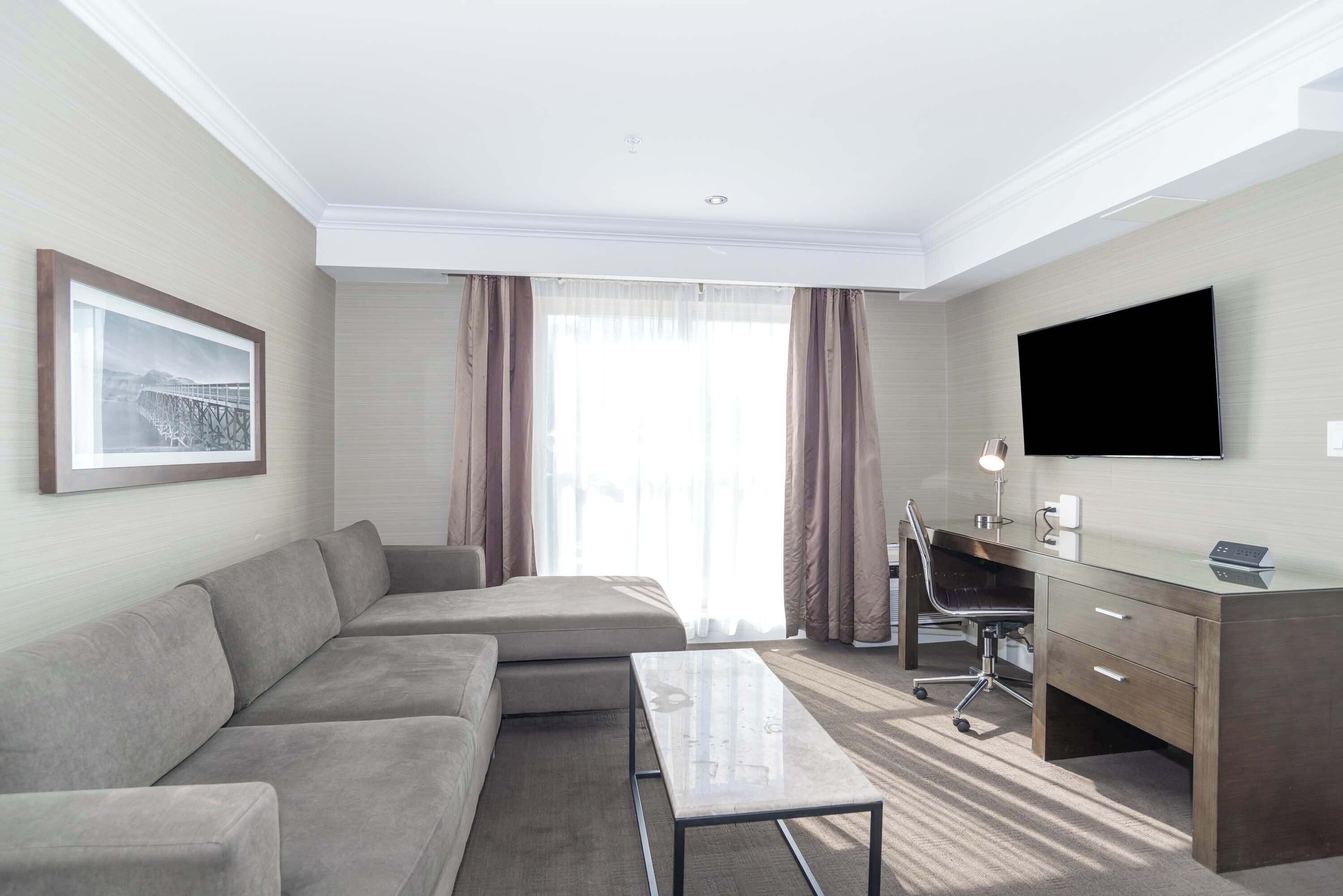 Quarto de hotel moderno com sofá, escrivaninha, televisão de tela plana e quadro na parede.