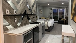 Cozinha moderna com micro-ondas embutido, pia e bancada para refeições rápidas. Há uma área de estar com sofá e poltrona, e uma janela grande com cortinas.