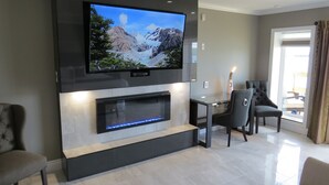 Uma sala de estar moderna com uma TV de tela plana acima de uma lareira, uma área de jantar com mesa e cadeiras, e uma grande janela com cortinas.