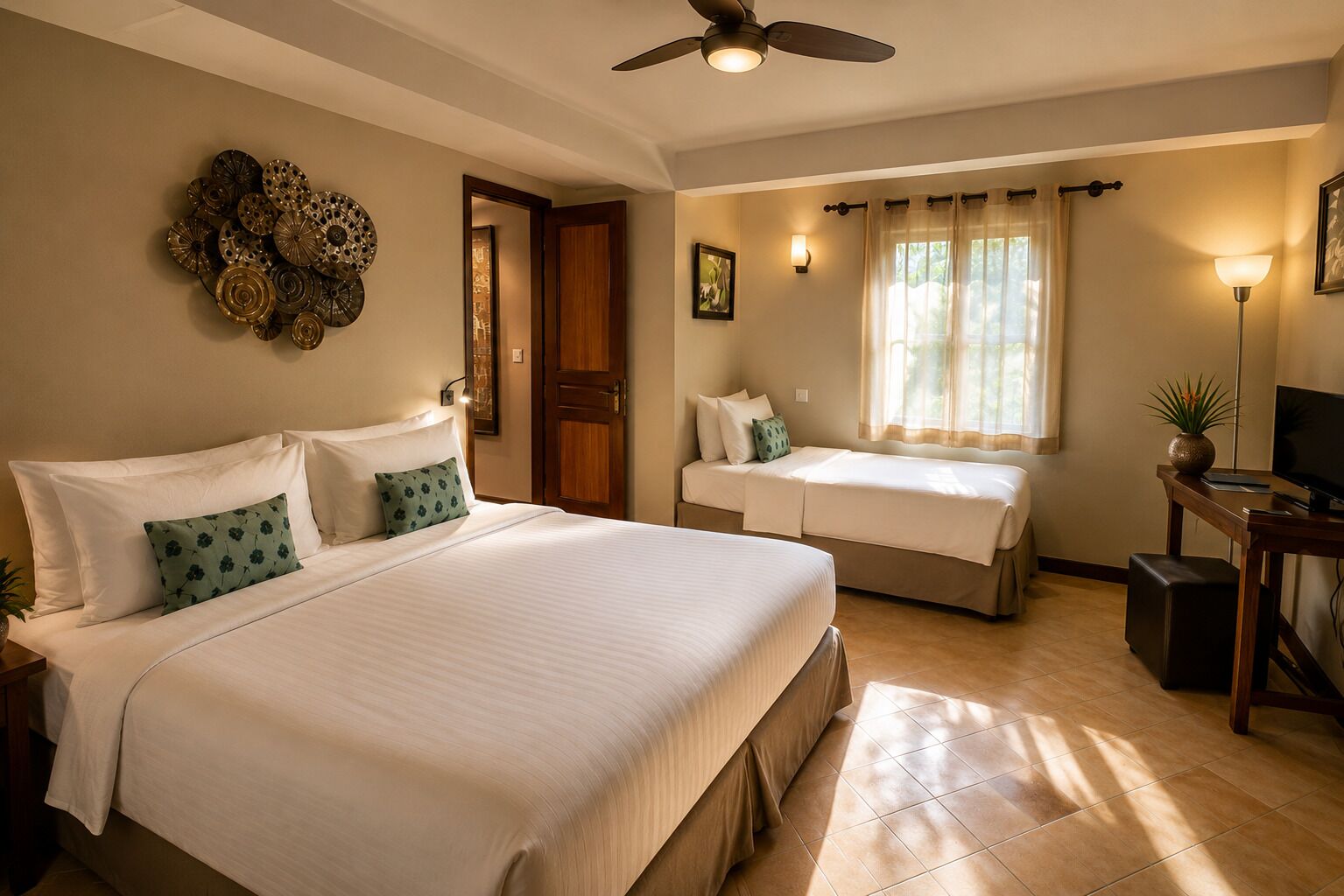 Quarto de hotel com uma cama grande, uma cama menor, uma mesa de trabalho com uma TV, um ventilador de teto e um detalhe decorativo na parede.