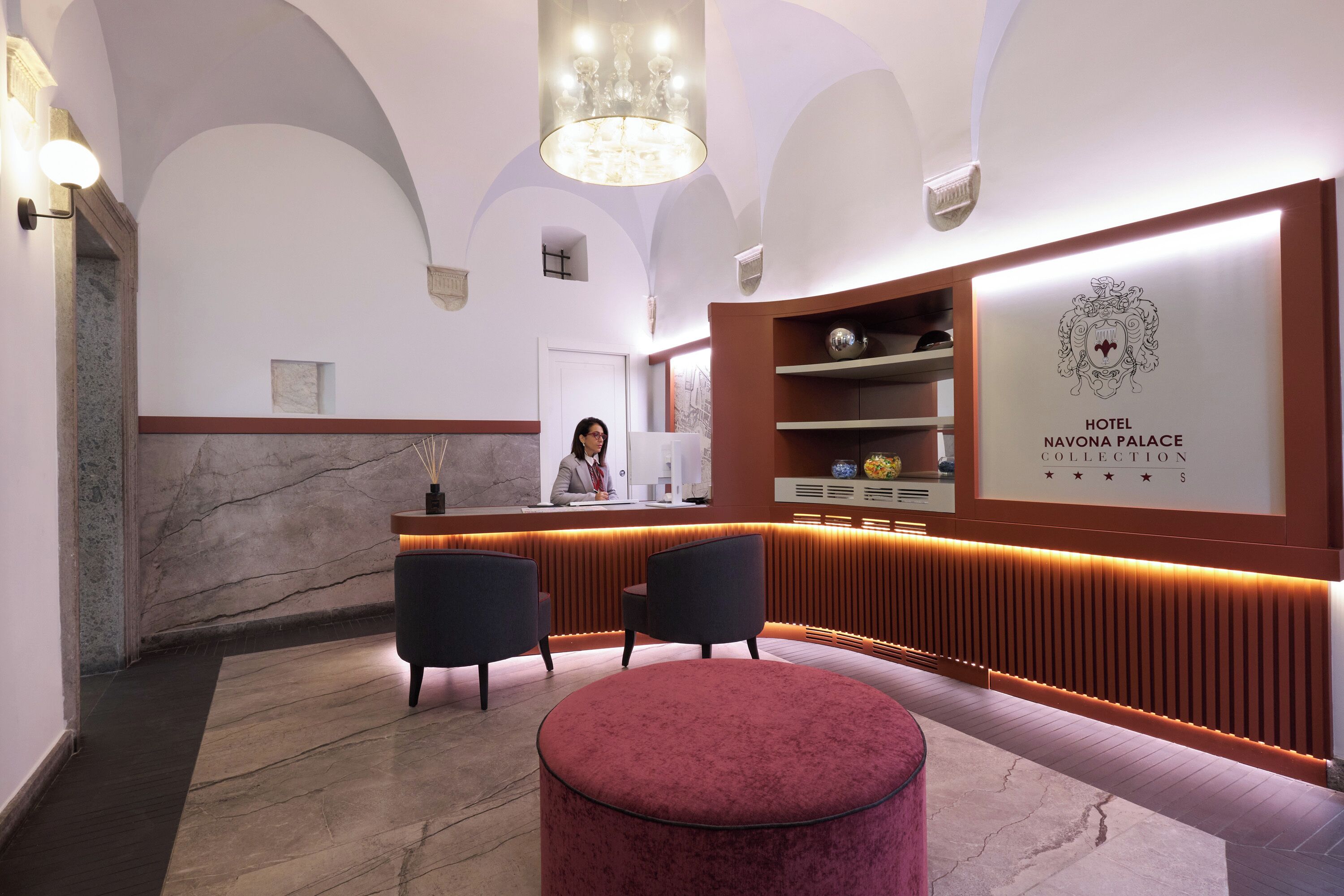 Uma área de recepção de hotel com piso de mármore, balcão de recepção, duas cadeiras, um banquinho vermelho e um candelabro.