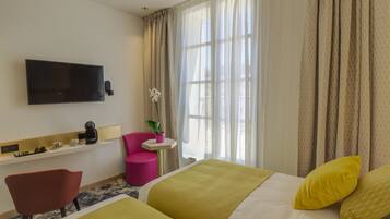Quarto de hotel com uma cama, televisão, secretária com cafeteira, cadeira e janela com cortinas.
