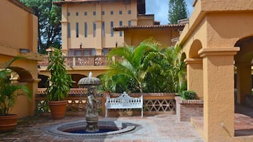 Um pátio com uma fonte, plantas em vasos, um banco branco e um edifício com varanda.