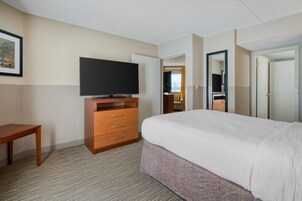 Quarto de hotel com cama, televisão sobre cômoda de madeira, mesinha e espelho.